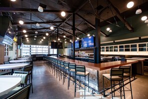 Um amplo espaço de bar com diversas opções de assento, incluindo banquetas e mesas, e teto alto com vigas aparentes e luminárias pendentes.