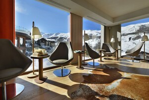 Quarto moderno com uma grande janela que oferece vista para montanhas cobertas de neve. Há várias cadeiras contemporâneas e uma pequena mesa com um livro.
