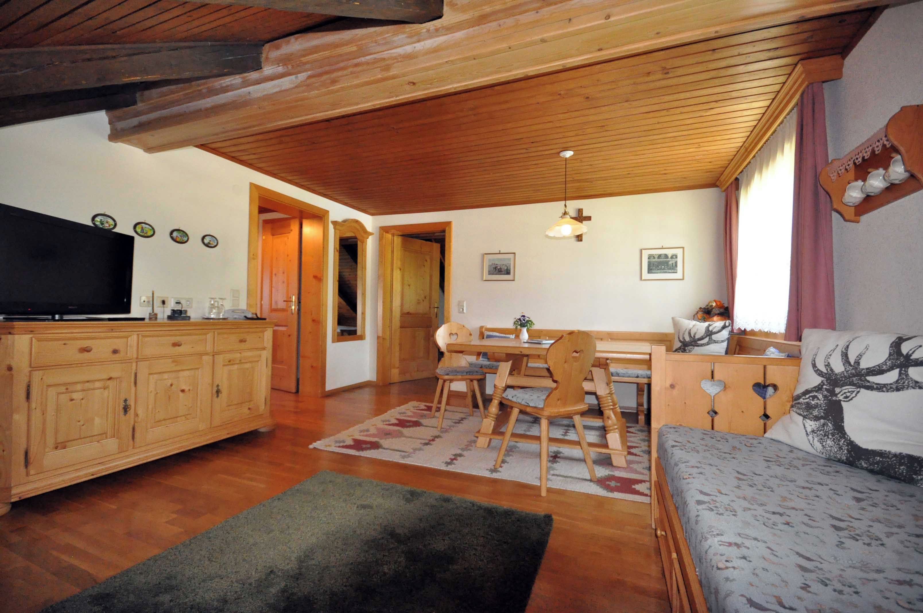 Uma sala de estar aconchegante com teto de madeira, televisão, área de jantar com mesa e cadeiras, e um sofá com almofada decorativa.