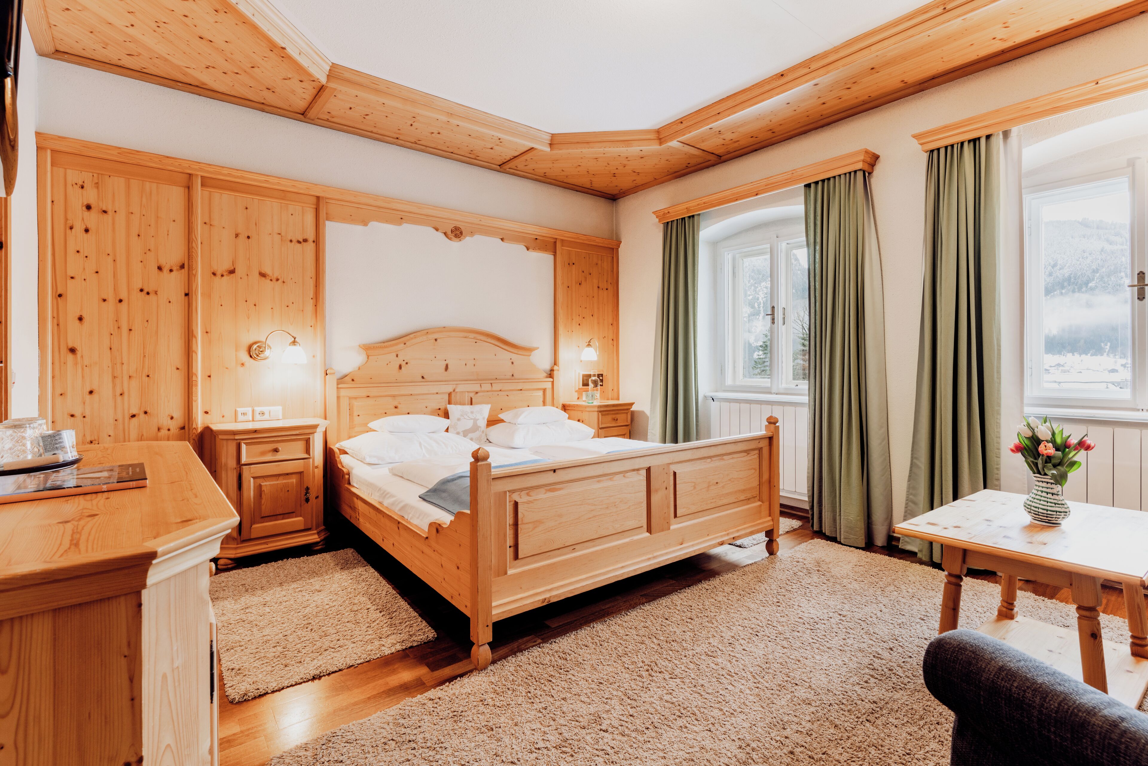 Quarto de madeira com cama, mesas de cabeceira, cortinas numa janela e vista para uma paisagem nevada.