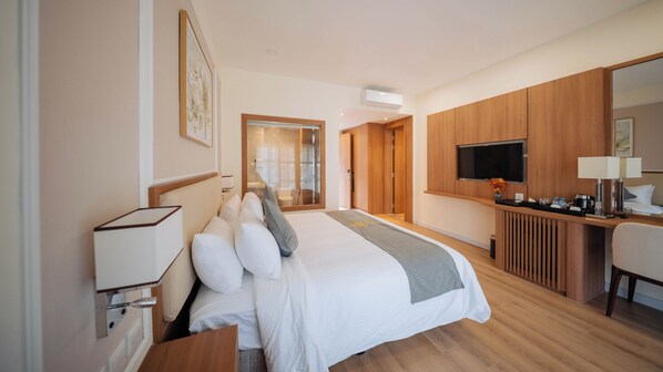 Quarto de hotel moderno com uma cama grande, uma escrivaninha com televisão de tela plana, um guarda-roupa de madeira e um banheiro visível através de uma porta de vidro.