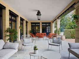 Uma área de estar coberta ao ar livre, com cadeiras, mesas, janelas amplas e plantas em vasos.
