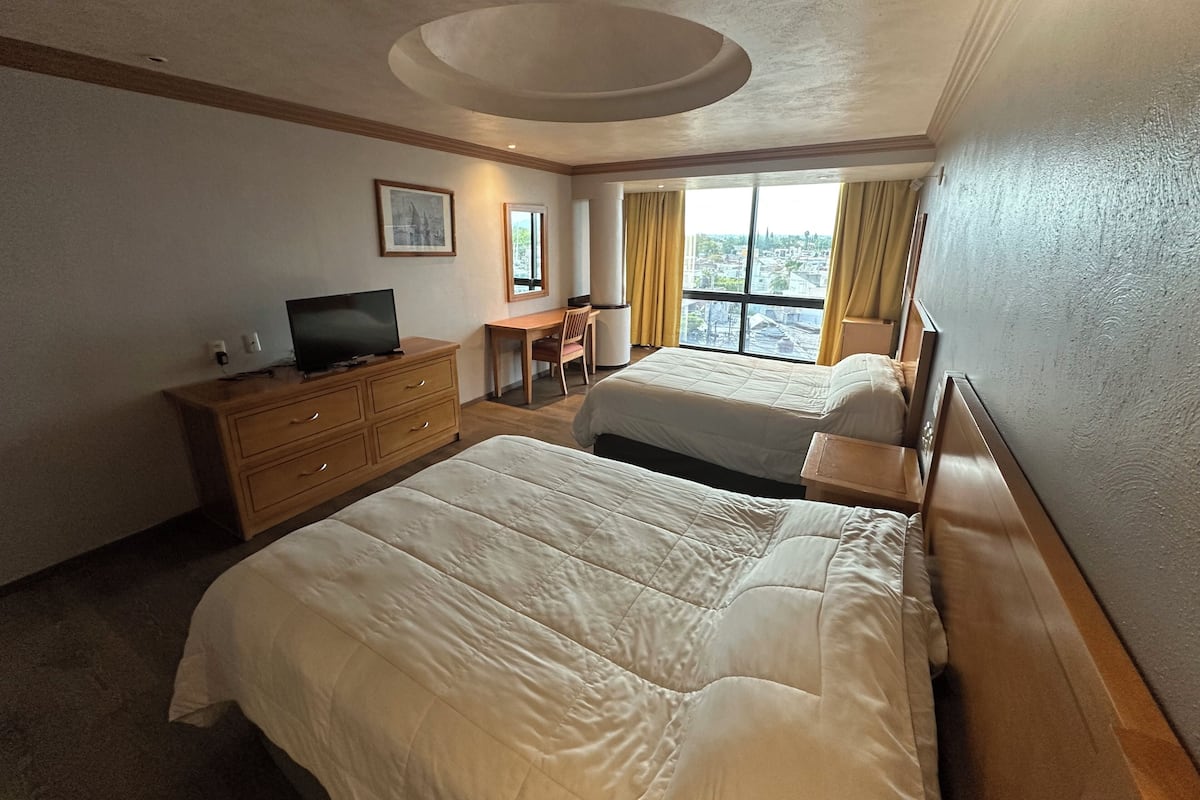 Quarto de hotel com duas camas, uma mesa de trabalho com cadeira, televisão e uma janela grande com cortinas.