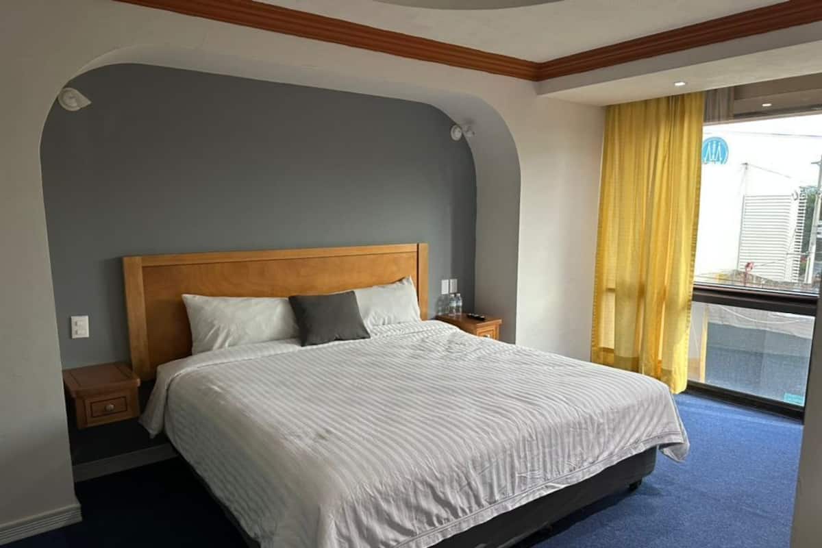 Quarto de hotel com uma cama grande, cabeceira de madeira, duas mesas de cabeceira e vista para a paisagem urbana através de uma janela com cortinas amarelas.