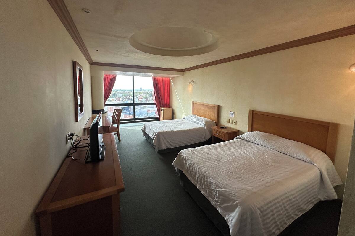 Quarto de hotel com duas camas, uma escrivaninha e uma cadeira. Possui uma janela grande com cortinas e vista para prédios.
