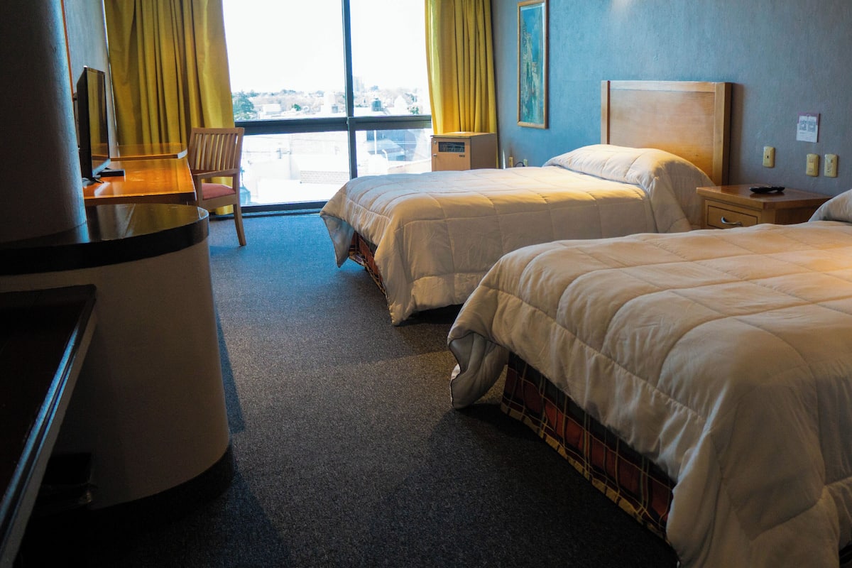Quarto de hotel com duas camas, uma escrivaninha, uma cadeira e uma janela grande com cortinas.