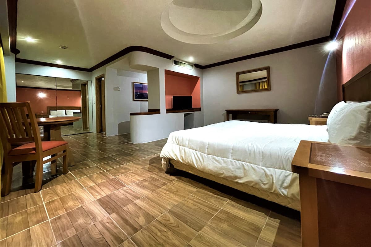 Quarto de hotel com uma cama grande, uma cadeira de madeira, uma escrivaninha e uma mesinha.