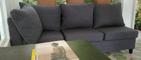 Um sofá cinzento com almofadas castanhas, uma mesa de centro verde com uma pilha de livros e vista para o verde lá fora.