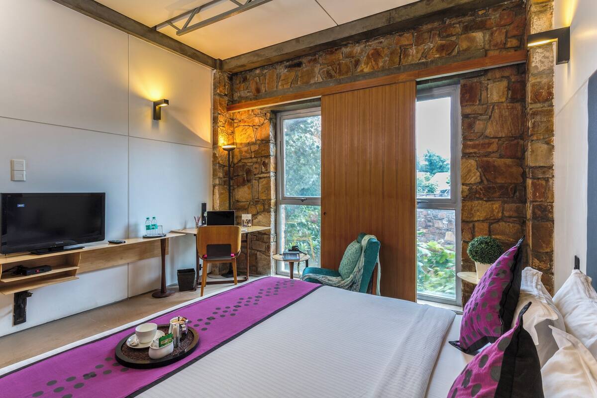 Quarto de hotel moderno com parede de detalhes em pedra, uma cama grande com almofadas, uma televisão, uma mesa de trabalho com cadeira e uma janela com vista para vegetação.