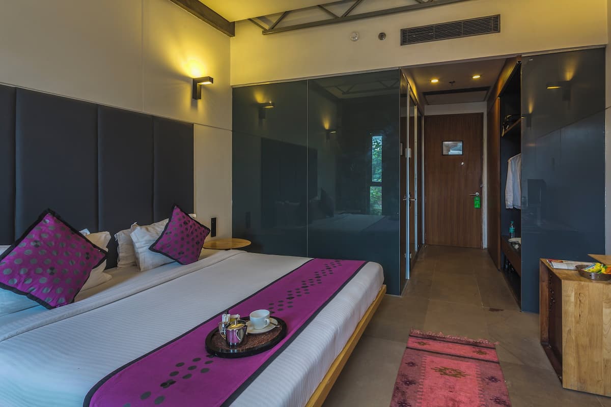 Quarto de hotel moderno com uma cama grande, divisória de vidro, porta de madeira e uma pequena mesa de cabeceira de madeira.