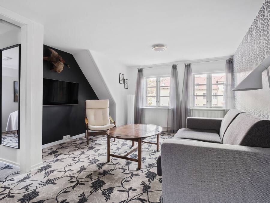 Uma sala de estar moderna com sofá, poltrona e uma pequena mesa de madeira. Uma parede preta com um elemento decorativo e uma janela com cortinas são visíveis.