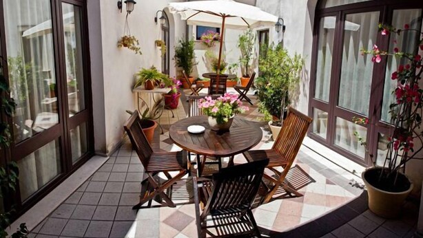 Um pátio com uma mesa de madeira redonda, quatro cadeiras e um guarda-sol branco. Há plantas em vasos e uma cesta suspensa com flores.
