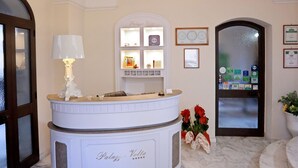 Uma área de recepção com um balcão branco, um candelabro, um tapete vermelho estampado e uma porta de vidro com painel fosco.
