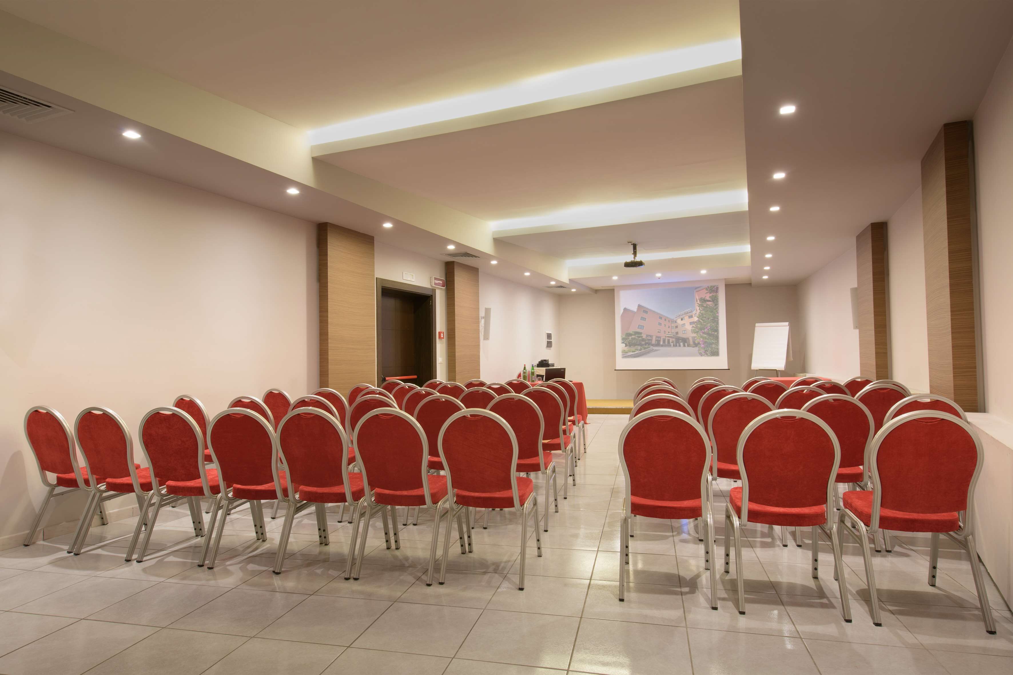 Uma sala de conferências com fileiras de cadeiras vermelhas, uma tela de projetor e uma área para apresentações.