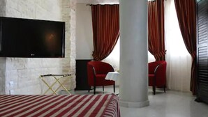 Quarto de hotel com cama, televisão na parede, duas cadeiras vermelhas, mesa e um pilar grande.