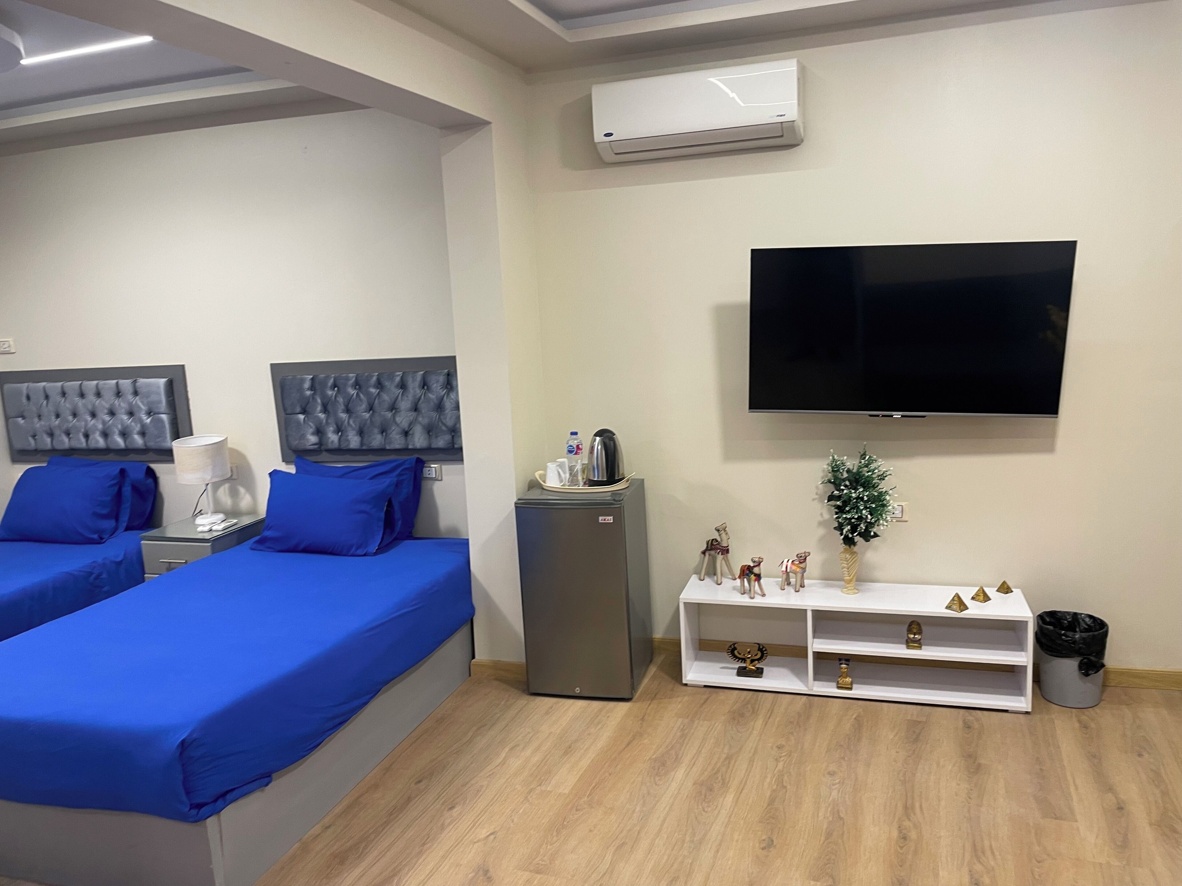 Quarto de hotel com duas camas, televisão, mini-frigorífico, mesa de cabeceira com abajur e prateleira decorativa com pequenas figuras.