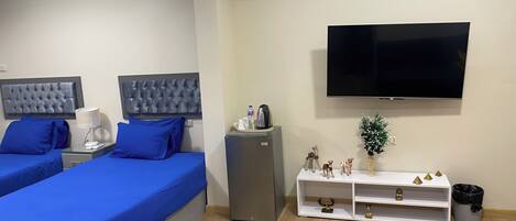 Quarto de hotel com duas camas, televisão, mini-frigorífico, mesa de cabeceira com abajur e prateleira decorativa com pequenas figuras.