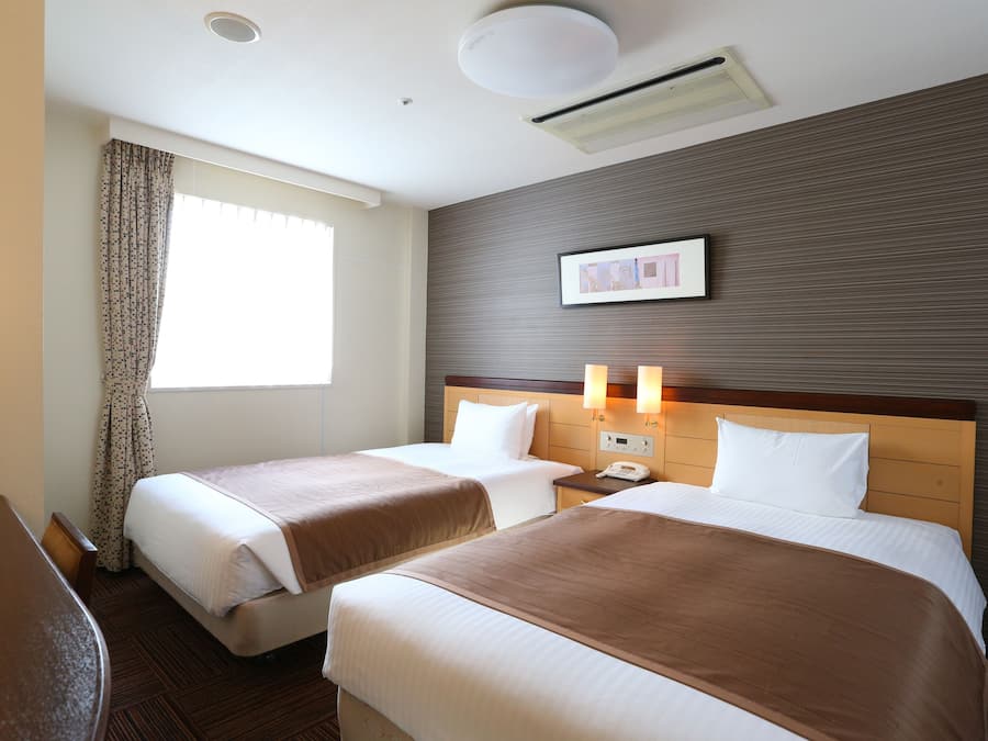 Quarto de hotel com duas camas, cabeceira de madeira, mesa de cabeceira com abajur, janela com cortinas e um quadro pendurado na parede.