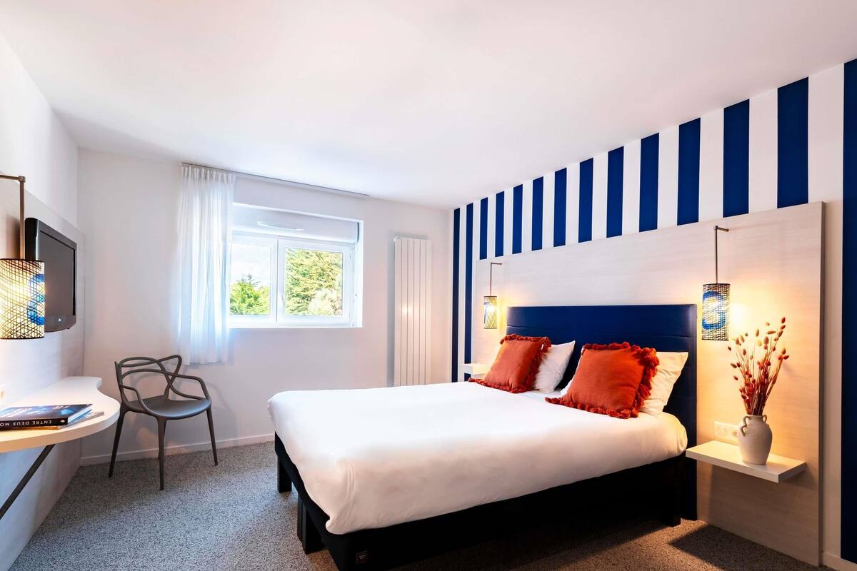 Quarto de hotel com cama, escrivaninha, cadeira, televisão, janela e parede com listras verticais.