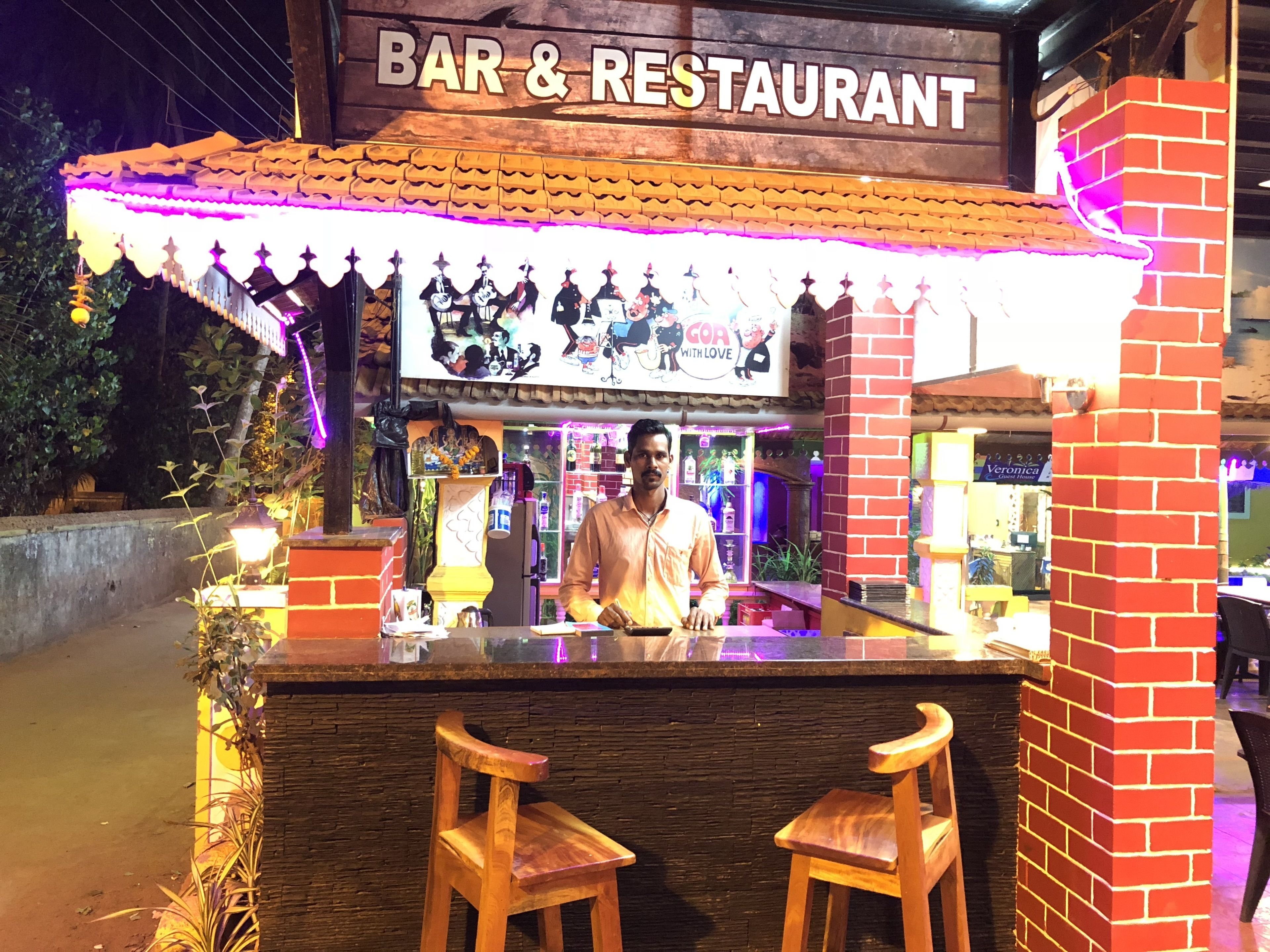 Um bar e restaurante com uma placa, balcão de madeira, dois bancos de madeira e uma pessoa atrás do balcão.