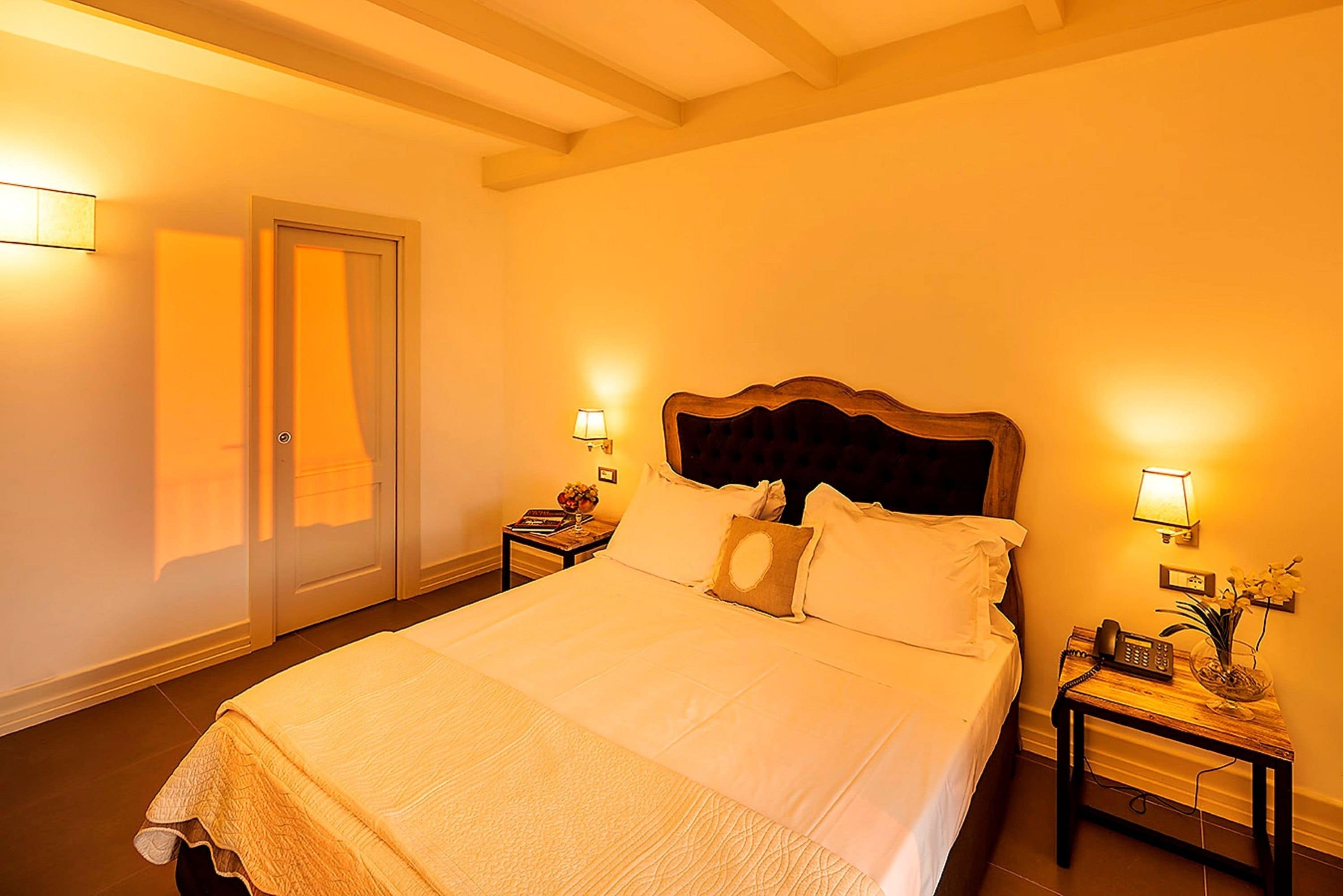 Uma cama bem arrumada com cabeceira de madeira, duas mesas de cabeceira com luminárias e uma porta com cortina.