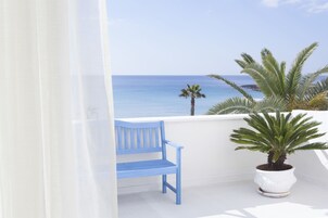 Varanda com cadeira azul e planta em vaso, com vista para o mar azul e palmeiras.