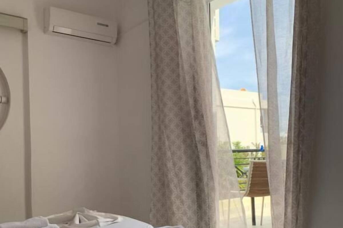 Quarto com cama, roupa de cama branca, janela com cortinas finas e ar condicionado na parede.