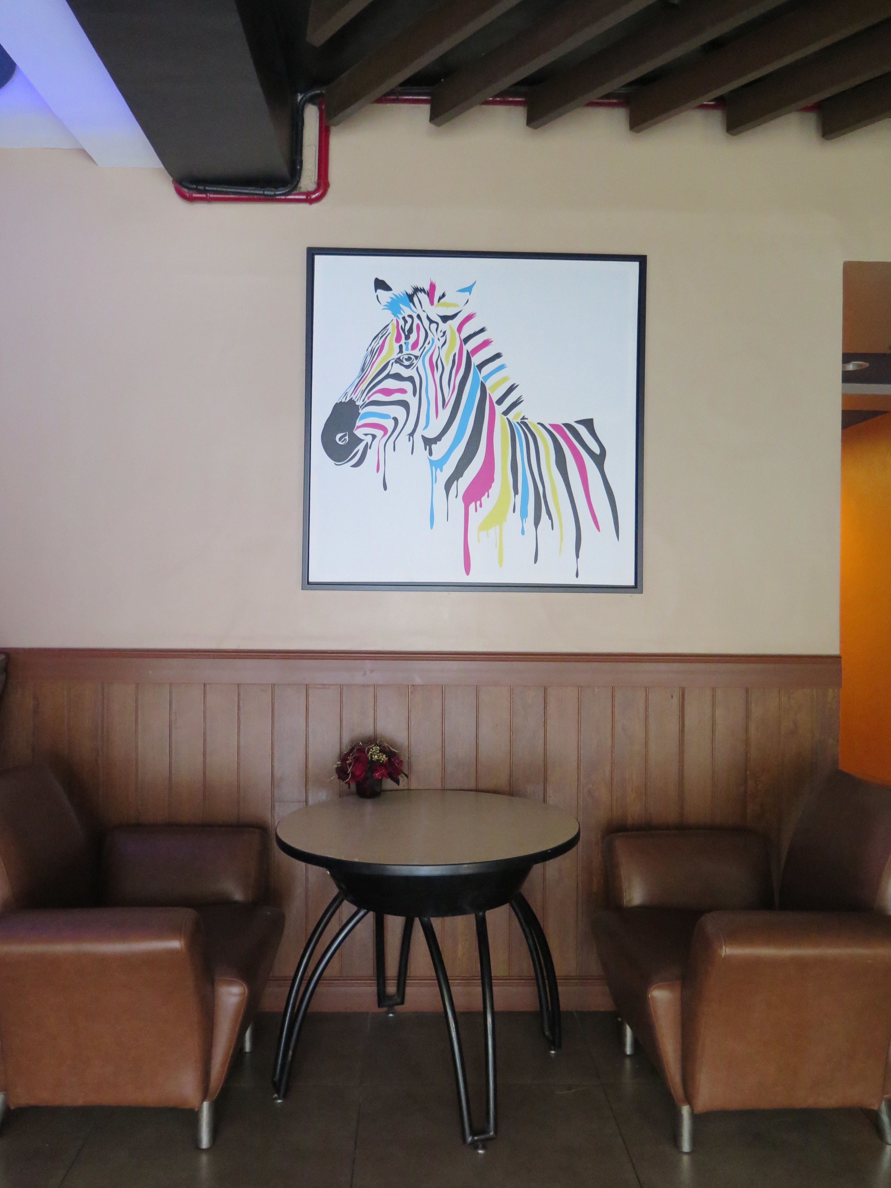 Um quadro de zebra fixado na parede, duas poltronas de couro marrom, uma pequena mesa com um vaso de flores em um quarto com painéis de madeira e piso de azulejos.