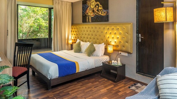 Quarto de hotel com uma cama grande, uma escrivaninha, uma cadeira, uma mesinha de cabeceira e um quadro na parede.