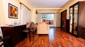 Quarto de hotel espaçoso com piso de madeira, sofá, escrivaninha com cadeira e vista para a cidade através de amplas janelas.