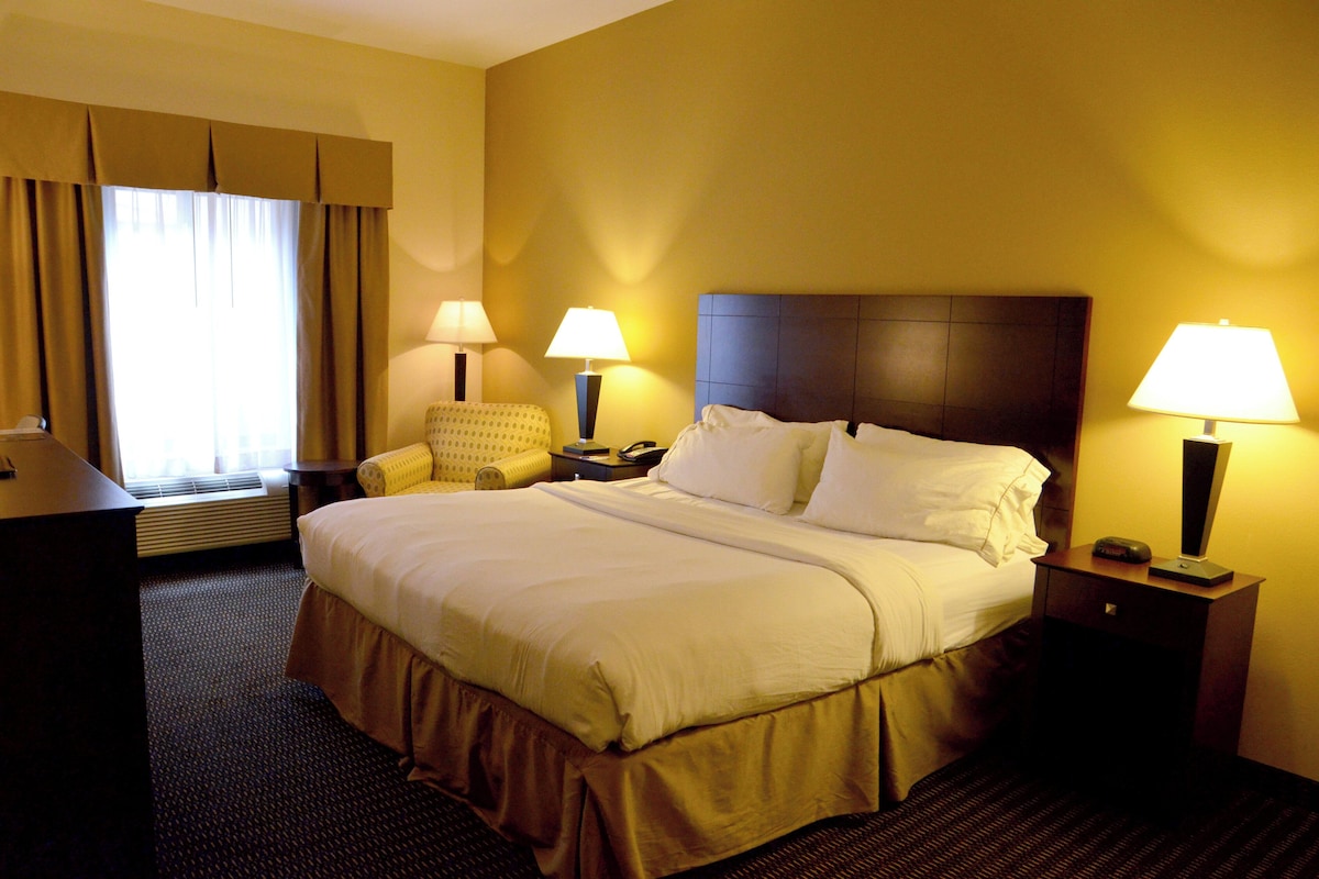 Quarto de hotel com uma cama grande, duas luminárias de cabeceira, uma cadeira, uma escrivaninha e uma janela com cortinas.