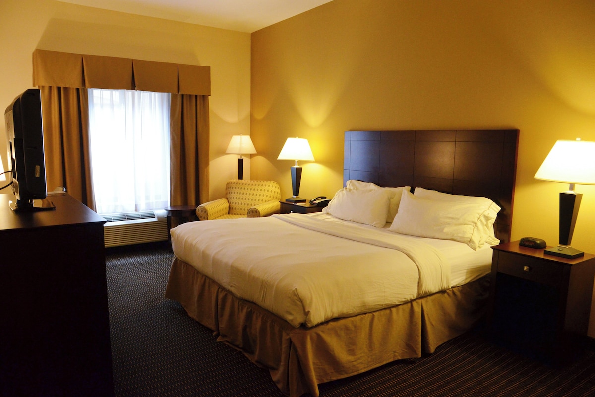 Quarto de hotel com uma cama grande, duas luminárias de cabeceira, uma cadeira, uma escrivaninha e uma janela com cortinas.
