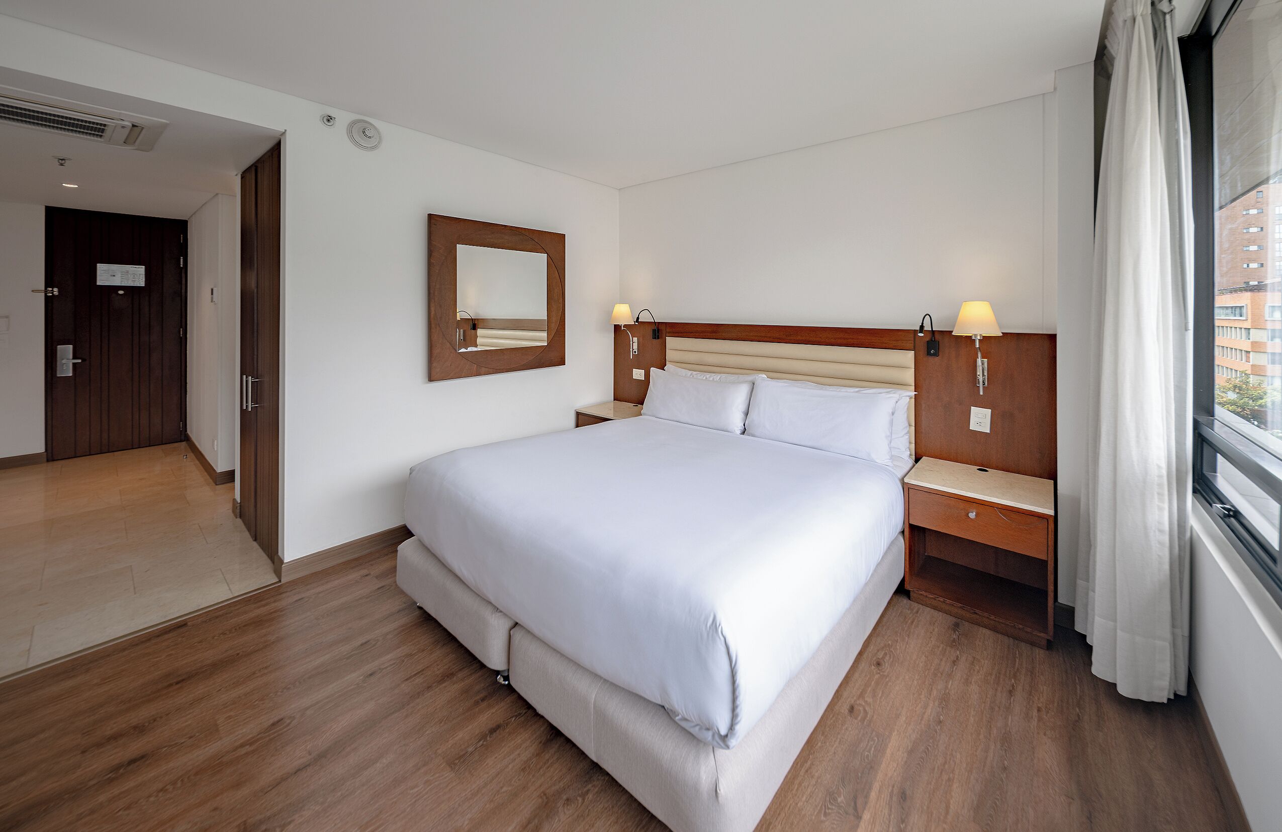 Quarto de hotel com cama grande, cabeceira de madeira, mesas de cabeceira e vista para edifícios pela janela.