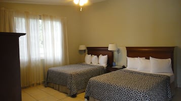 Quarto de hotel com duas camas, um ventilador de teto, mesa de cabeceira com abajur e janela com cortinas.