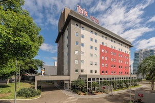 Um hotel com vários andares, com uma placa de destaque 'ibis Hotel', uma entrada coberta e uma área ajardinada com árvores e arbustos.