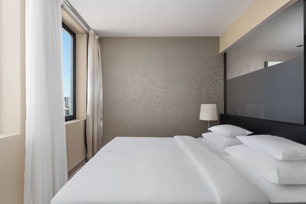 Quarto de hotel com uma cama grande, mesa de cabeceira, luminária e vista para a cidade pela janela.