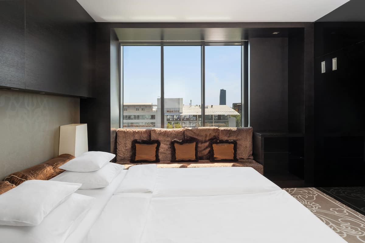 Quarto de hotel moderno com uma cama grande, vista para a cidade através de uma janela ampla e uma área de estar aconchegante com almofadas.