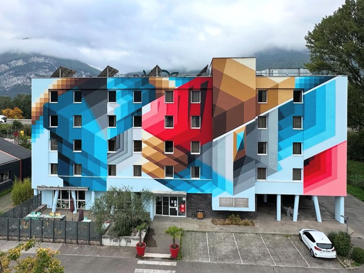 Um edifício moderno com fachada geométrica, área de estacionamento e montanhas ao fundo.
