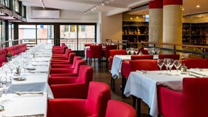 Um restaurante com fileiras de cadeiras vermelhas e mesas brancas, janelas amplas e vista para a cidade.