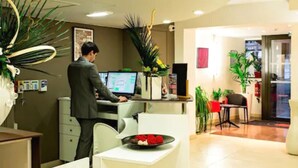 Um homem trabalha numa mesa com um computador, cercado por plantas e uma área de recepção com uma cadeira vermelha e uma mesa branca.