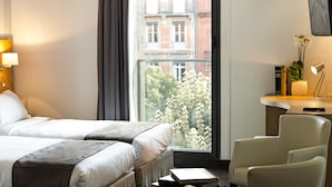 Quarto de hotel com duas camas, uma escrivaninha com uma luminária, uma cadeira e vista para um edifício e árvores pela janela.