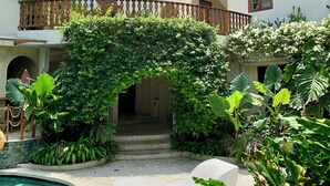 Um edifício branco com janelas e varandas em arco, rodeado por vegetação exuberante e uma piscina.