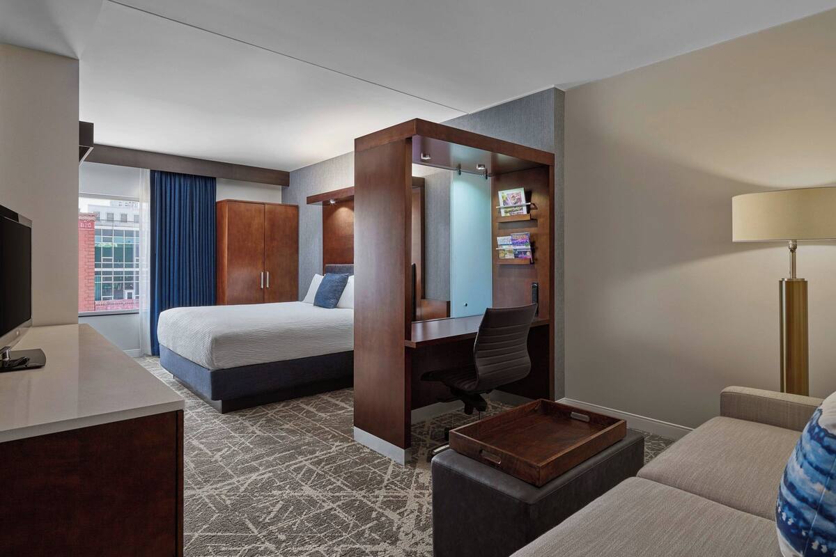 Quarto de hotel com cama, escrivaninha, cadeira e televisão.