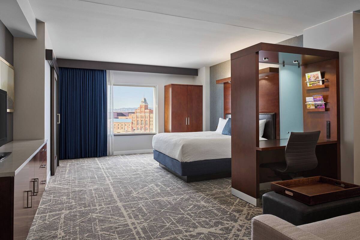 Quarto de hotel com uma cama grande, uma escrivaninha com cadeira, uma televisão e vista para a paisagem urbana.