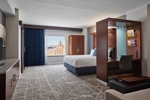 Quarto de hotel com uma cama grande, uma escrivaninha com cadeira, uma televisão e vista para a paisagem urbana.