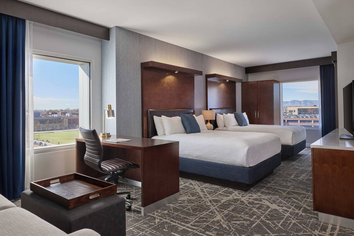 Quarto de hotel com uma cama grande, uma mesa e uma cadeira. Há uma janela com vista para edifícios e montanhas.