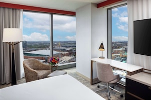 Quarto de hotel com uma janela grande, uma cama, uma escrivaninha, uma cadeira e uma televisão.
