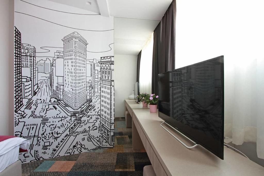 Quarto de hotel moderno com uma televisão de tela grande na parede, um mural com vista urbana, uma pequena planta em vaso e um tapete estampado.