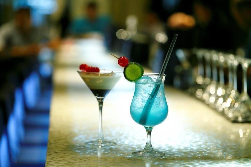 Dois cocktails sobre um balcão de bar, com pessoas desfocadas ao fundo.