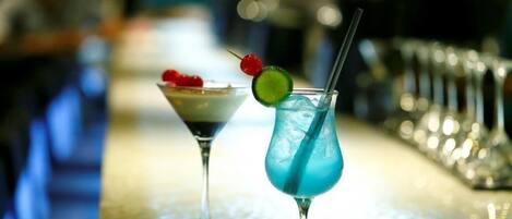 Dois cocktails sobre um balcão de bar, com pessoas desfocadas ao fundo.